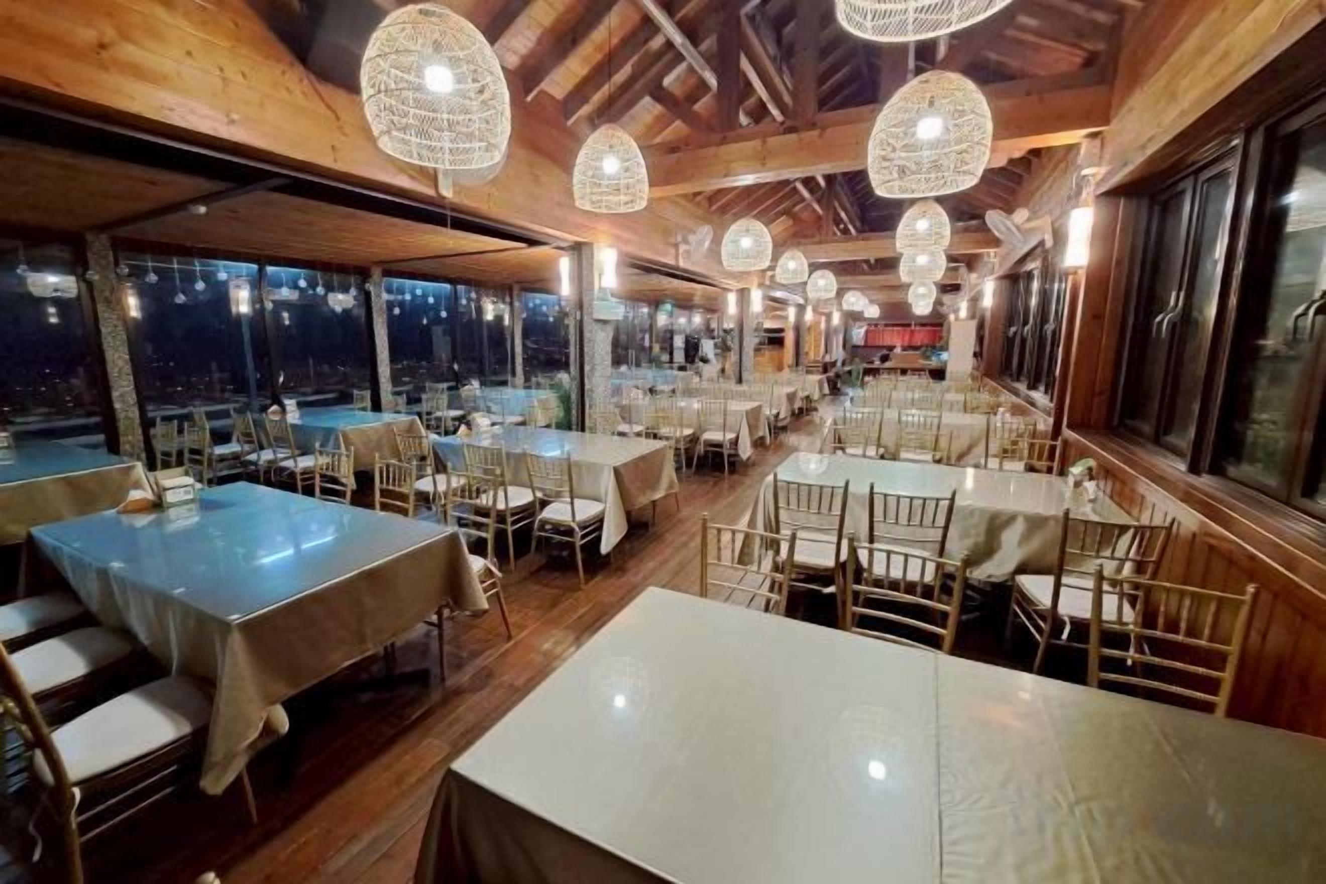 Espaço de refeições interno com mesas, cadeiras, vigas de madeira e luminárias pendentes.