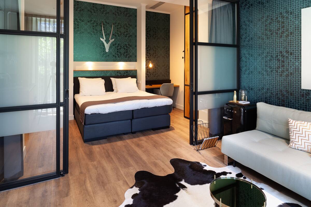 Quarto de hotel moderno com uma cama grande, um sofá, uma escrivaninha e vista através de uma porta de vidro.