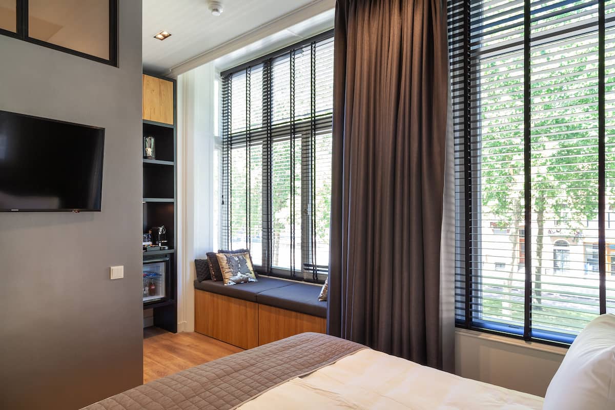 Quarto com cama, um banco embutido com assentos, televisão montada na parede e janelas amplas com persianas.