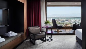Quarto de hotel moderno com uma janela grande, televisão de tela plana, sofá, poltrona, uma mesinha com uma planta e uma cama.