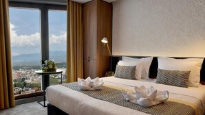 Quarto de hotel com uma cama grande, uma mesa de cabeceira, uma luminária e vista da cidade através de grandes janelas.