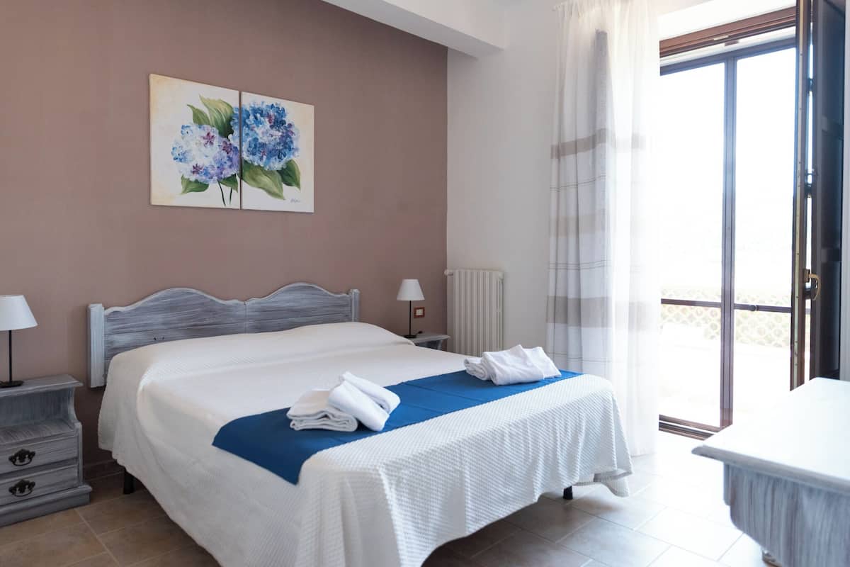 Cama bem arrumada com detalhes em azul, mesa de cabeceira com abajur, radiador e vista para o exterior por uma porta de correr.