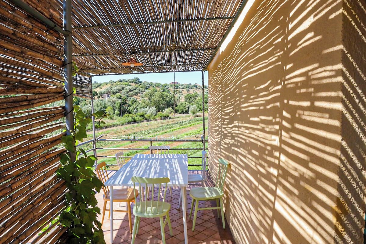 Um pátio externo com mesa e cadeiras, paredes de bambu e vista para uma paisagem verde.