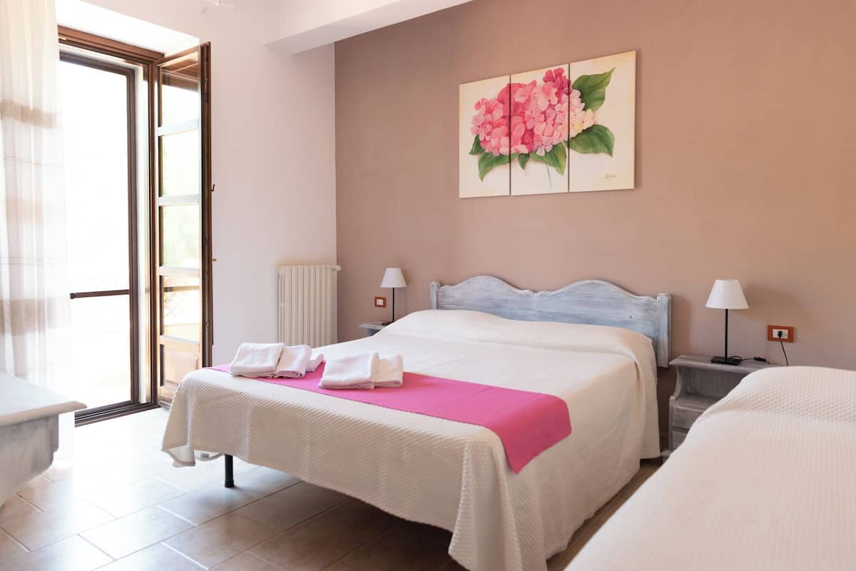 Quarto de hotel com uma cama, mesas de cabeceira, um radiador e um quadro de flores na parede.