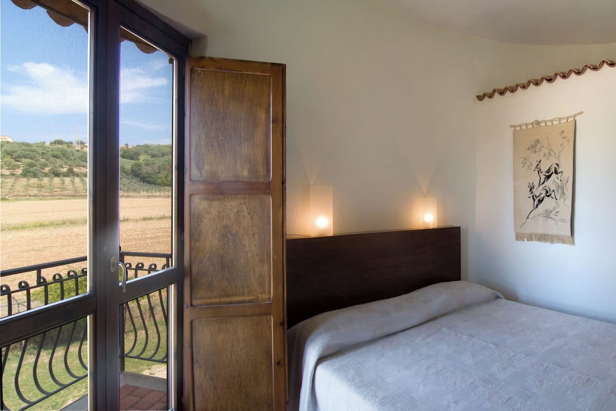 Quarto com uma cama grande, cabeceira de madeira, obra de arte na parede, banheiro com pia e espelho, e varanda com vista para o campo.