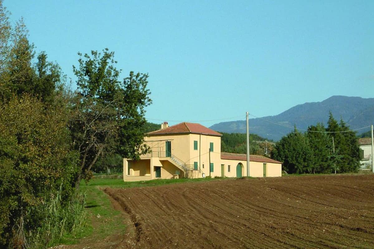 Uma casa de dois andares com telhado vermelho, localizada em zona rural, com um campo arado em primeiro plano e montanhas ao fundo.