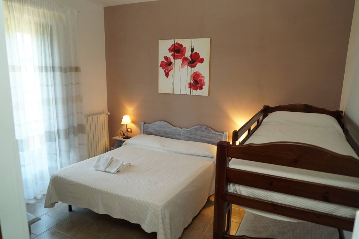 Quarto com uma cama, uma mesa de cabeceira com abajur, um radiador e um quadro de flores vermelhas na parede.