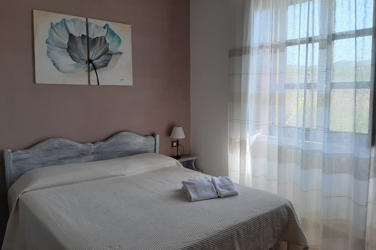 Cama bem arrumada com uma toalha dobrada, uma luminária de cabeceira e um quadro de uma flor na parede.