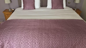 Uma cama bem arrumada com um edredom rosa texturizado, almofadas brancas e uma mesinha de cabeceira de madeira com um abajur.