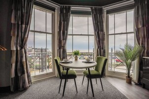 Quarto de canto com mesa redonda, duas cadeiras, planta em vaso e vista da cidade através de grandes janelas.