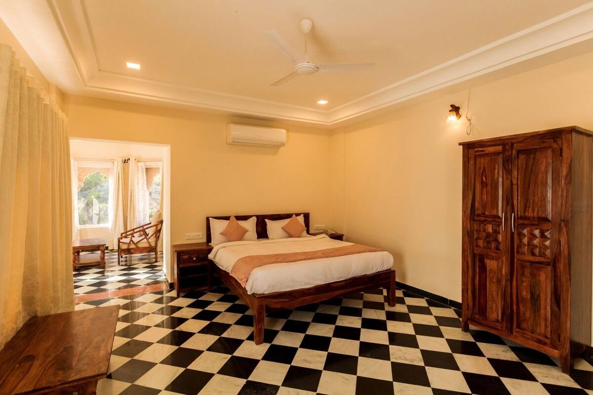 Quarto com piso xadrez, cama com travesseiros, guarda-roupa de madeira, mesa de cabeceira, cadeira e ventilador de teto.