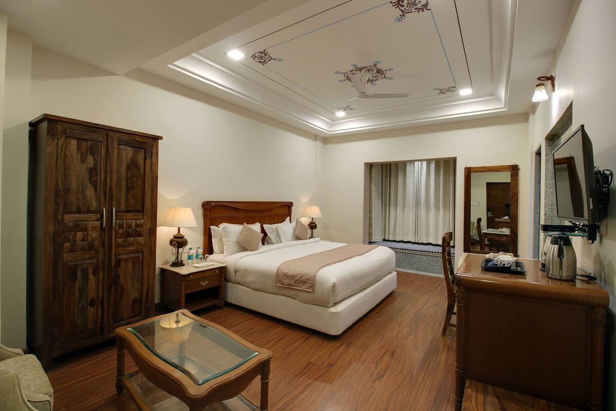 Quarto de hotel com guarda-roupa de madeira, cama com lençóis brancos, mesa de cabeceira com luminárias, pequena mesa de madeira e espelho na parede.