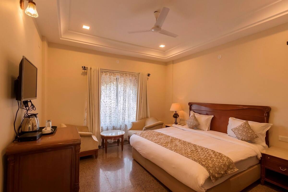 Quarto de hotel com uma cama, uma escrivaninha com cafeteira, uma cadeira, uma mesa, uma televisão e um ventilador de teto.