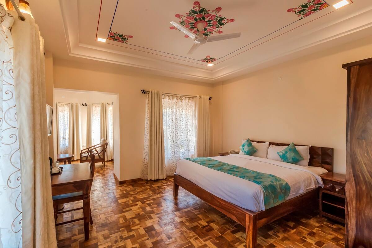 Quarto com piso de madeira, cama com colcha verde, escrivaninha com cadeira e ventilador de teto.