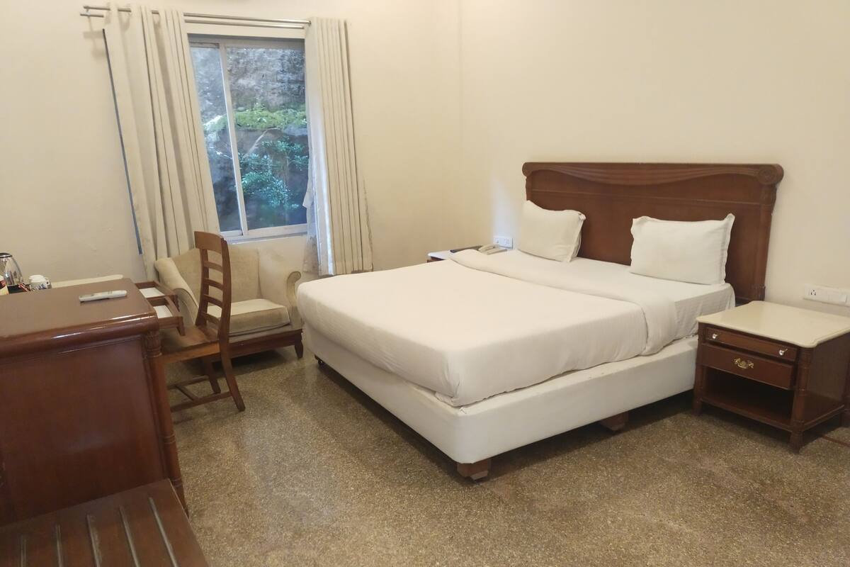 Quarto de hotel com cama, escrivaninha, cadeira, mesa de cabeceira e janela com cortinas.