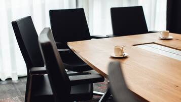 Uma mesa de conferência com quatro cadeiras pretas, uma chávena branca sobre a mesa e uma janela com cortinas ao fundo.