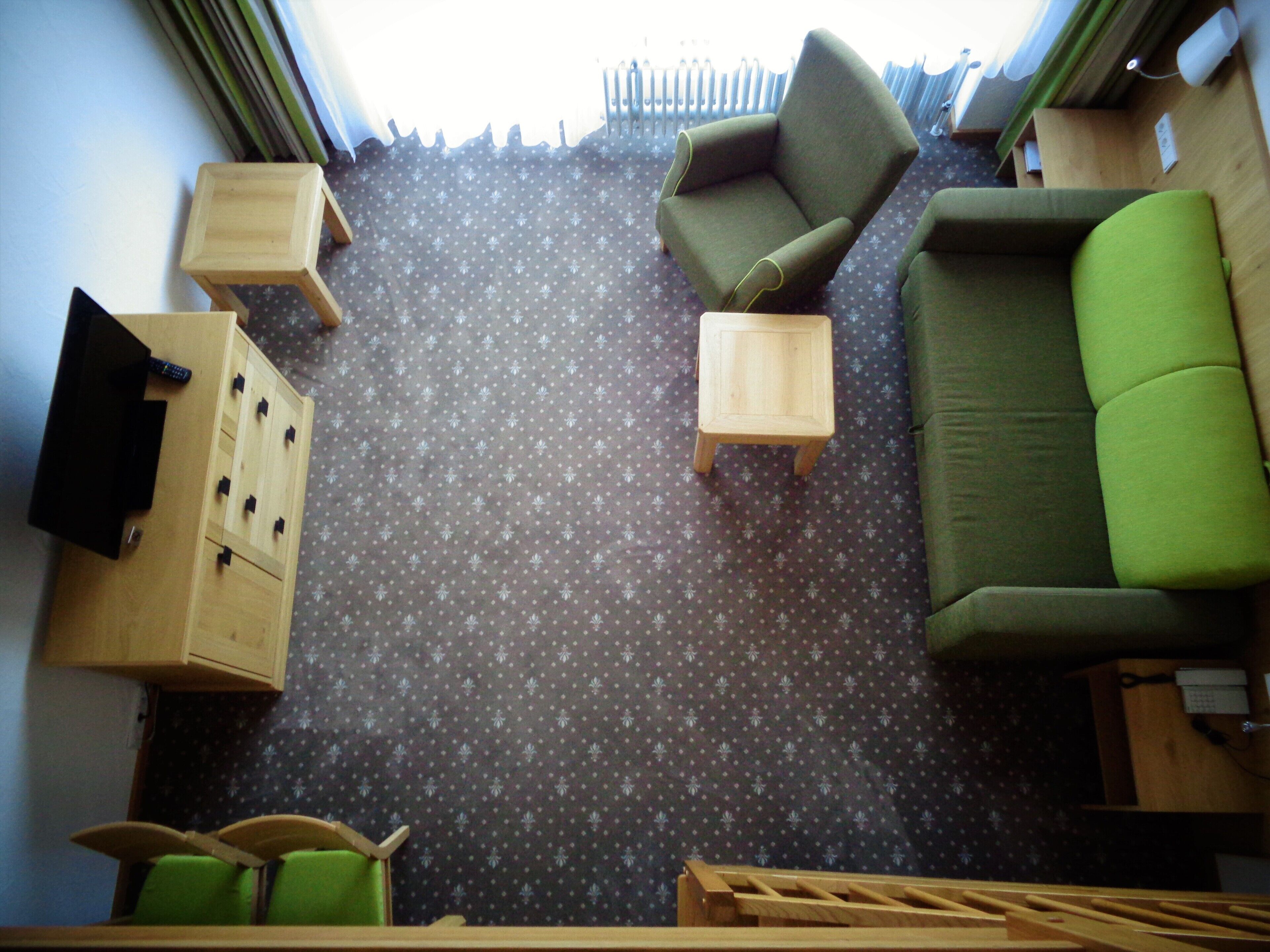 Quarto compacto com um sofá verde, uma pequena mesa de madeira e uma cadeira verde. Há um armário de madeira e um pequeno banquinho de madeira. O quarto possui um tapete estampado e uma janela com vista para uma cerca.