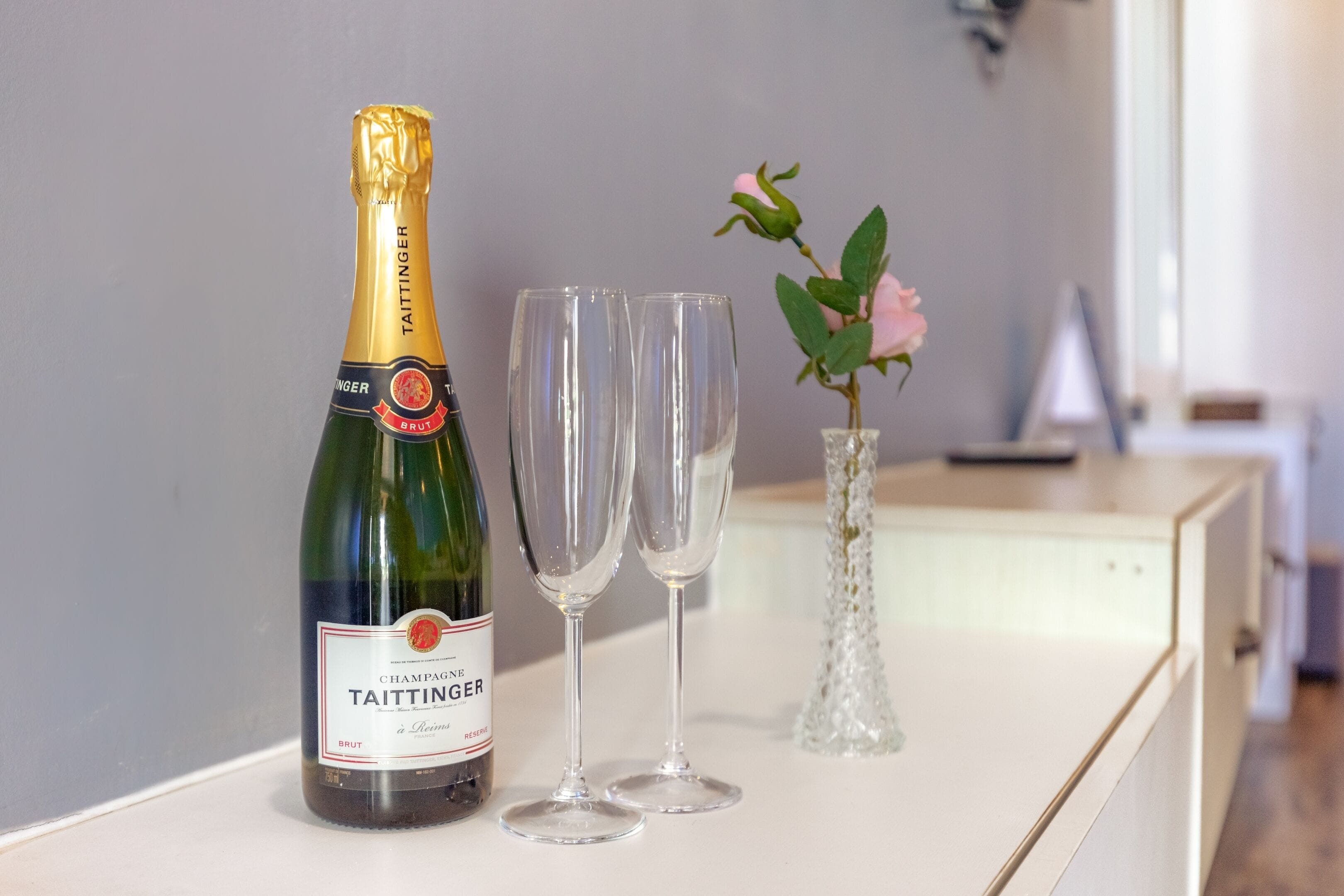 Uma garrafa de champanhe Taittinger e dois copos vazios sobre uma superfície branca.