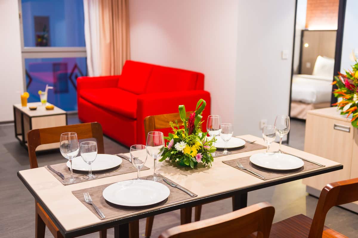 Uma sala de jantar com uma mesa posta para dois, um sofá vermelho ao fundo e um vaso de flores sobre a mesa.