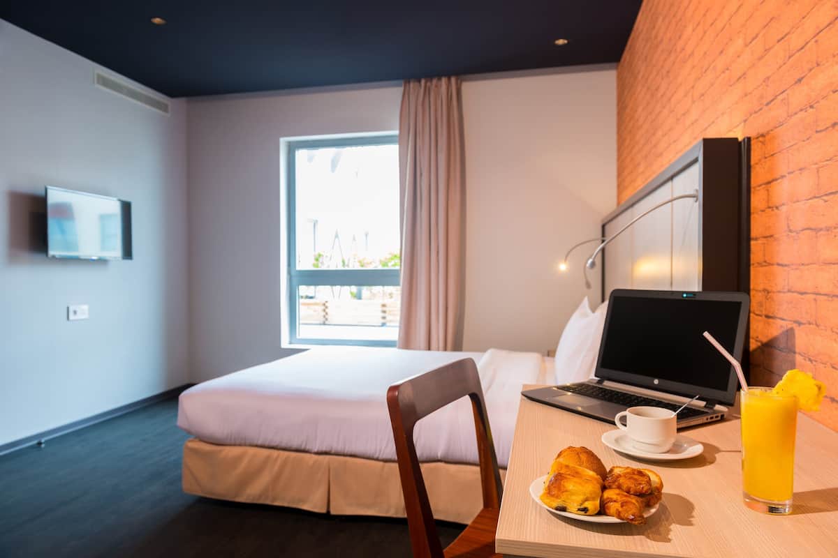 Quarto de hotel com cama, secretária com laptop, chávena de café, prato de pastelaria e copo de sumo de laranja.
