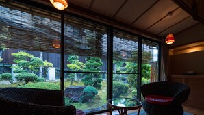 Quarto com uma grande janela com vista para um jardim, duas cadeiras de vime, uma mesa pequena e um lustre.