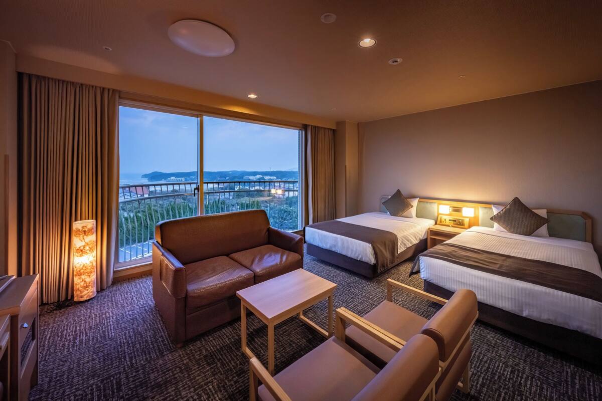 Quarto de hotel com duas camas, um sofá, uma mesinha e vista para uma ponte e horizonte urbano.