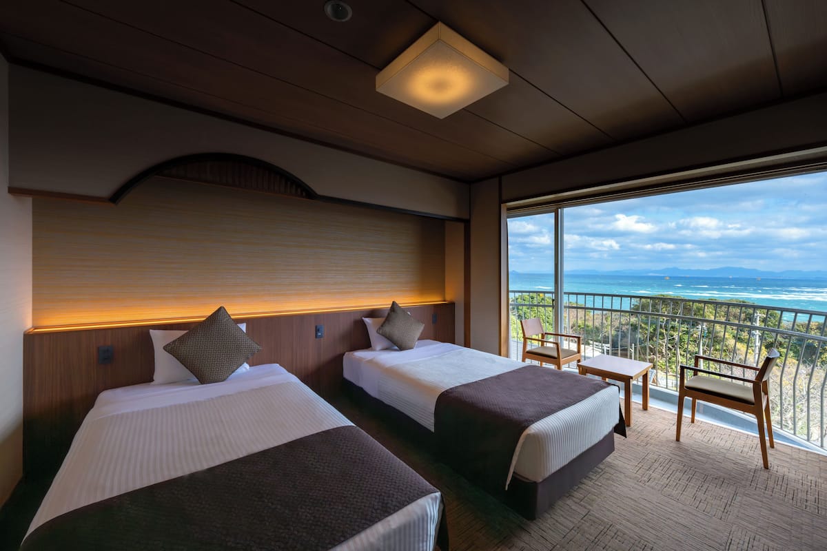 Quarto de hotel com duas camas, varanda e vista para o mar, além de uma pequena mesa e cadeira.