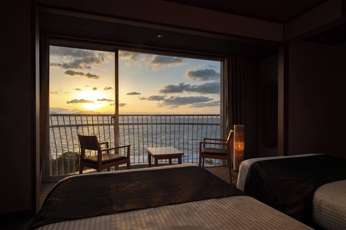 Quarto de hotel com uma grande janela com vista para o mar ao pôr do sol, duas camas, uma mesa pequena e duas cadeiras.