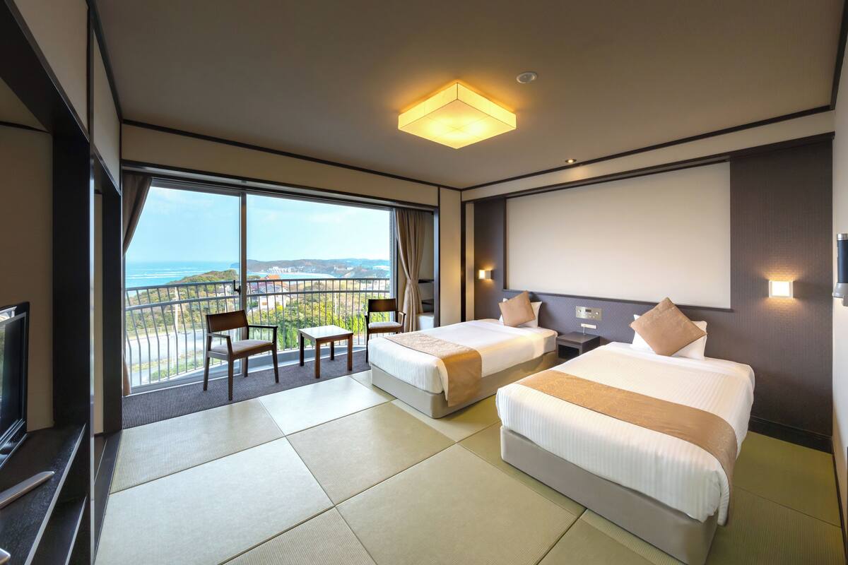 Quarto de hotel com duas camas, televisão, varanda com vista e uma janela grande.