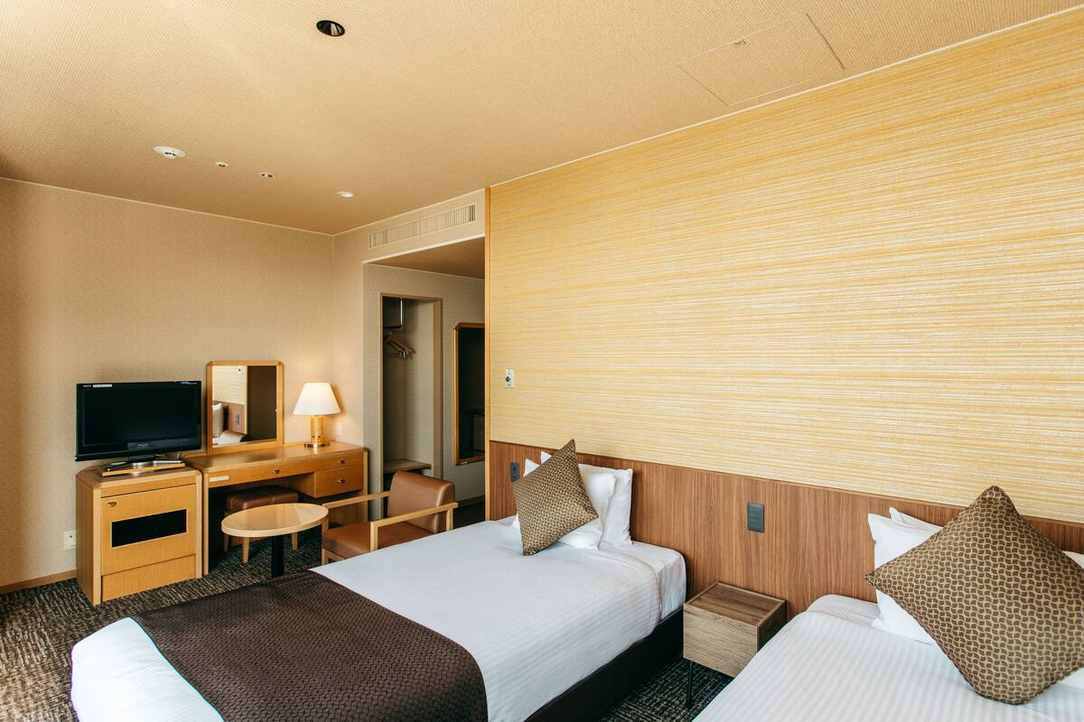 Quarto de hotel com duas camas, uma escrivaninha com televisão, uma cadeira e uma mesa pequena.