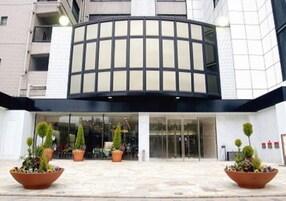 Um edifício moderno com entrada em vidro curvo, plantas em vasos e calçada de azulejos.