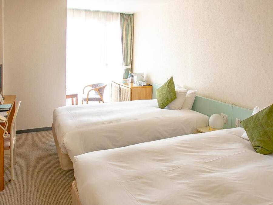 Quarto de hotel com duas camas, uma escrivaninha, uma cadeira e uma janela com cortinas.