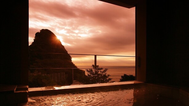 Varanda com vista para o oceano ao pôr do sol, piscina e montanha ao fundo.
