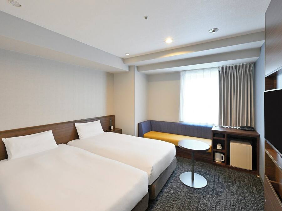 Quarto de hotel com duas camas, uma mesinha, um sofá, uma televisão e uma janela com cortinas.