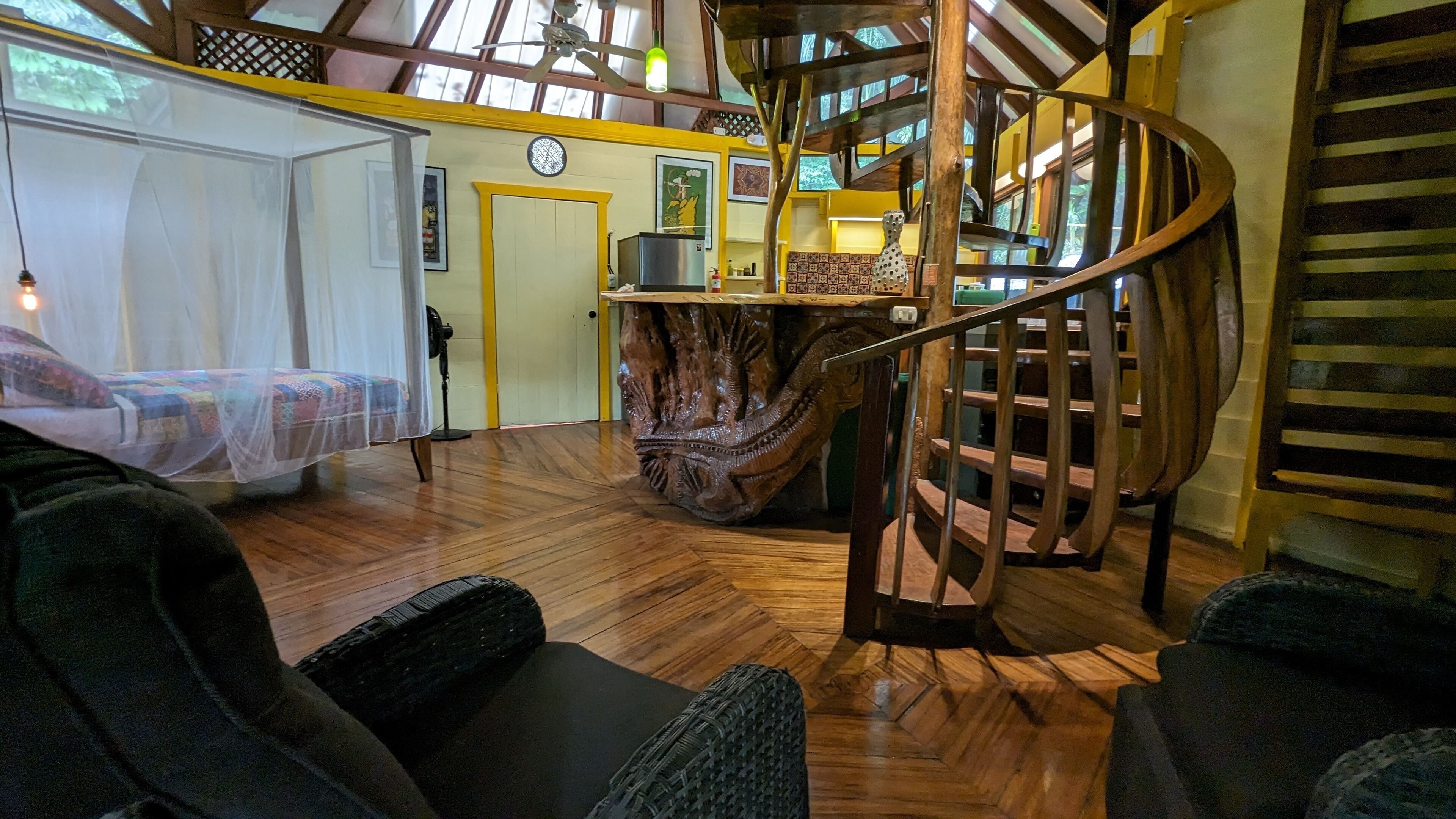 Uma escadaria em espiral com corrimãos de madeira leva a um andar superior. Uma cama com dossel branco e um edredom colorido é visível em primeiro plano.