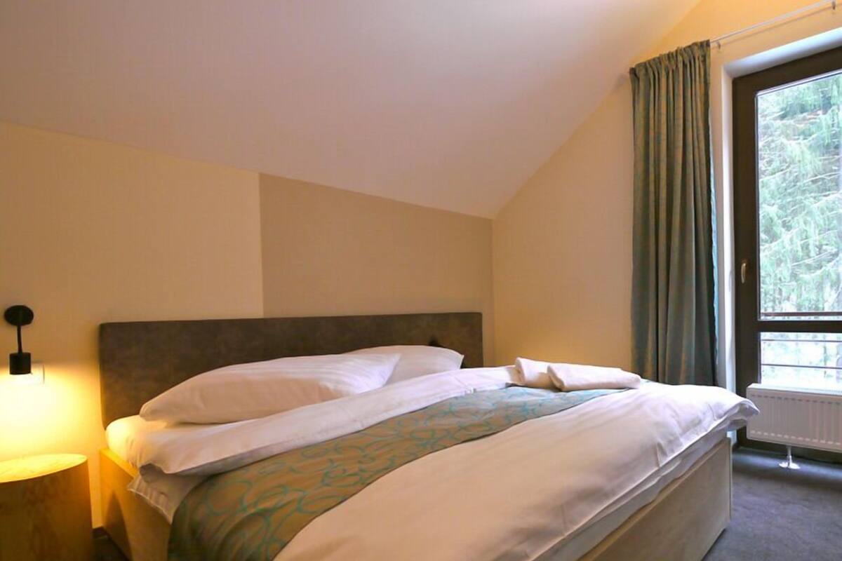 Quarto de hotel com cama, mesa de cabeceira, luminária e janela com cortinas.