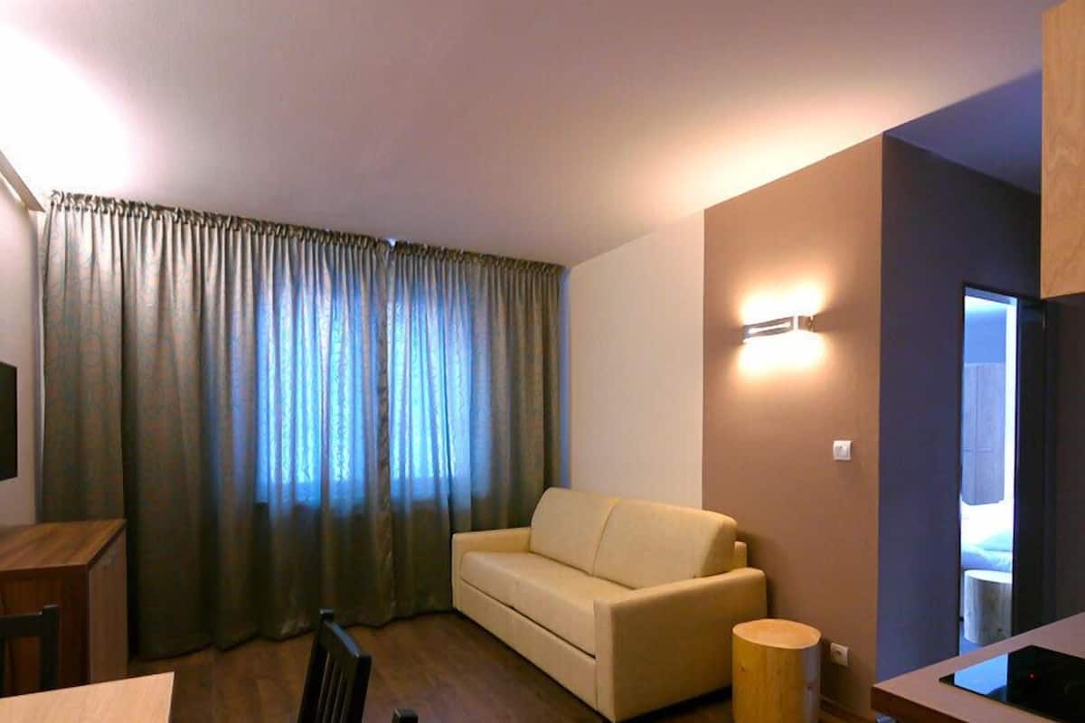 Um espaço de estar compacto com sofá, mesa de jantar, cadeiras, um banquinho de madeira e um aparelho de luz fixado na parede.