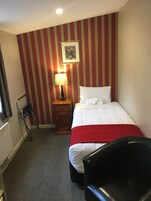 Quarto de hotel com parede listrada em vermelho e dourado, uma cama com roupa de cama branca, um cobertor vermelho, uma mesa de cabeceira com abajur e uma cadeira.