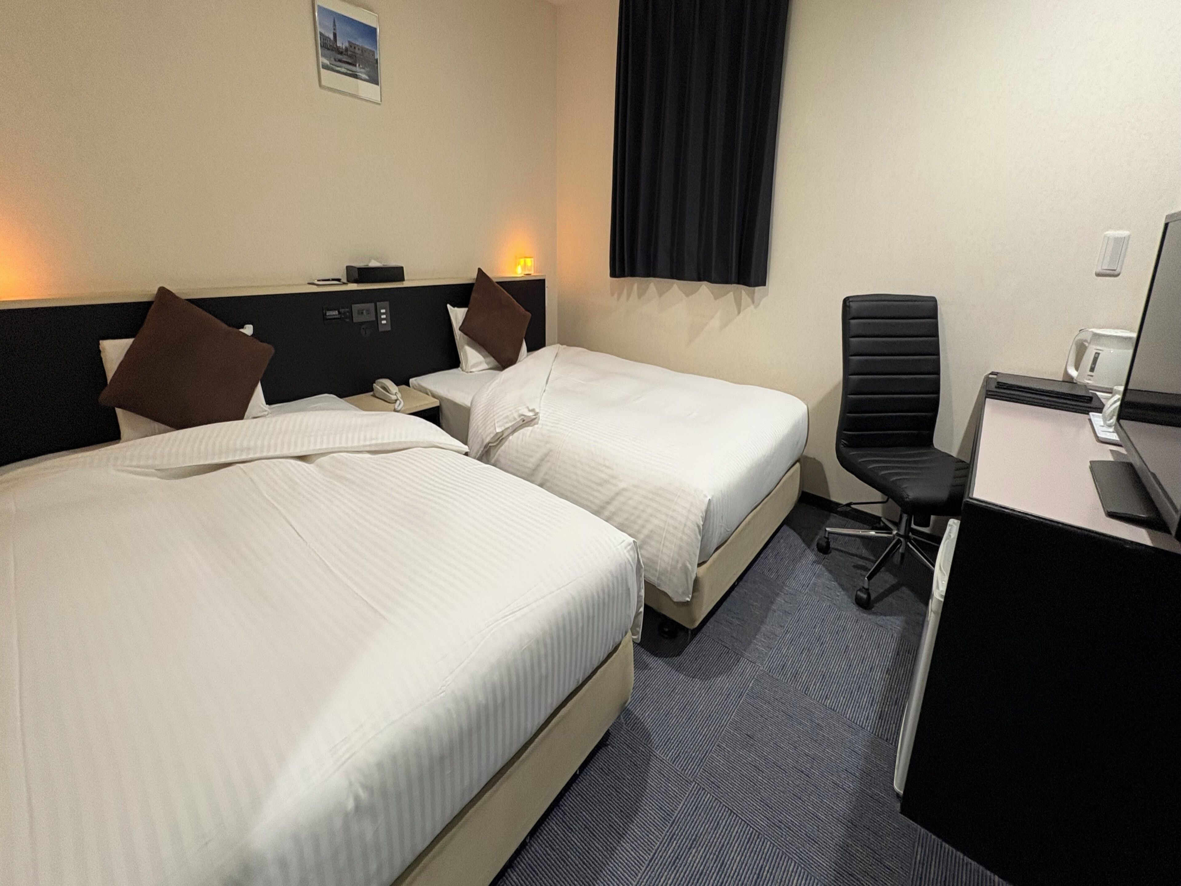 Quarto de hotel com duas camas, uma escrivaninha com computador, uma cadeira e uma pequena mesa com abajur.