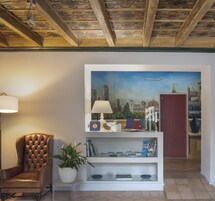 Quarto com pintura de uma grande paisagem urbana, uma poltrona de couro, uma planta em vaso e uma vitrine de vidro com livros e objetos decorativos.