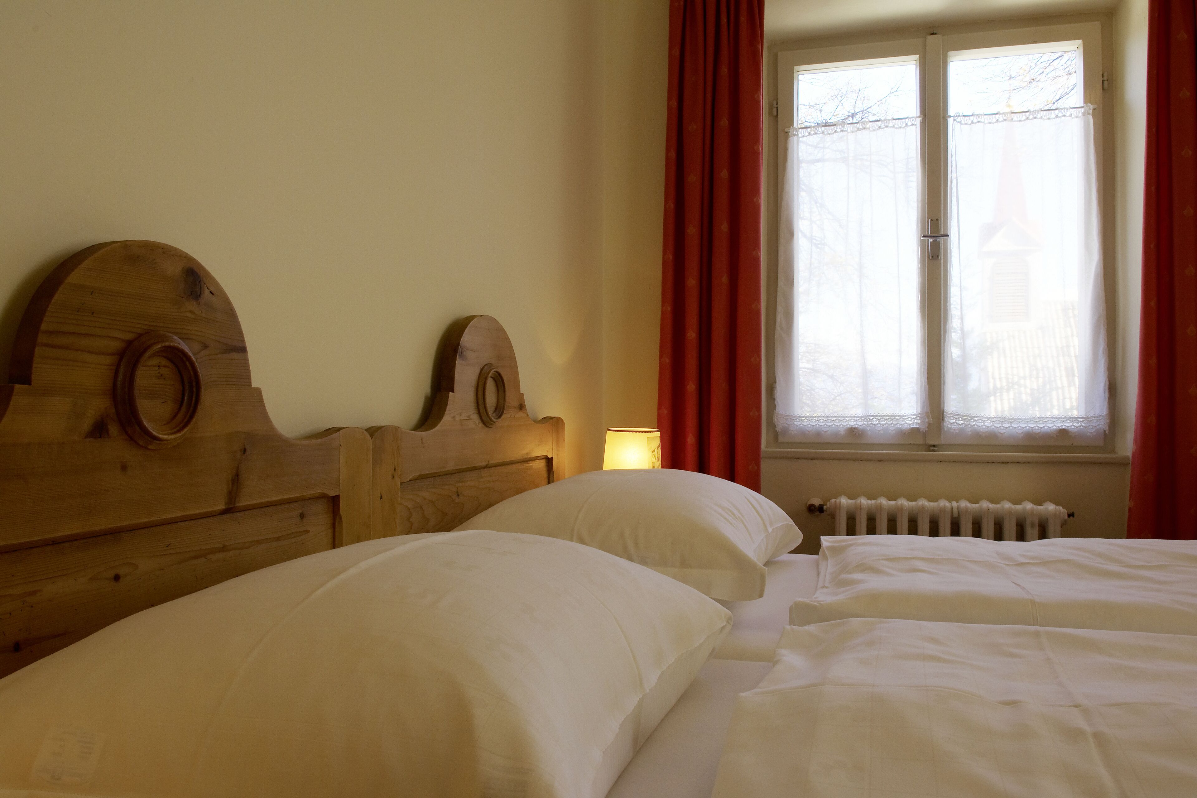 Quarto com duas camas, cabeceiras de madeira, janela com cortinas vermelhas e radiador.