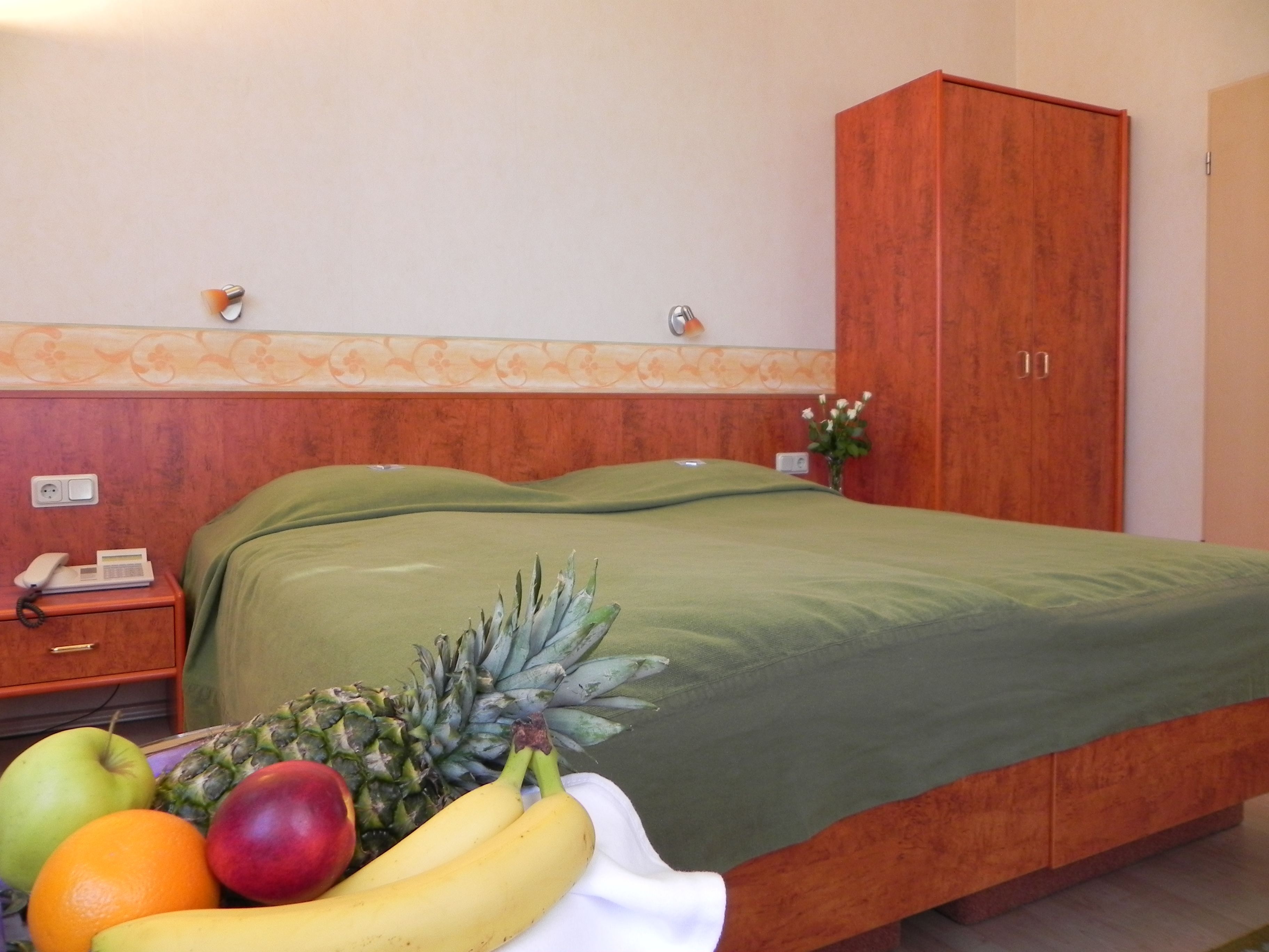 Quarto de hotel com uma cama, um guarda-roupa de madeira, uma mesa de cabeceira com telefone e uma fruteira com maçãs, laranjas, bananas e abacaxi.