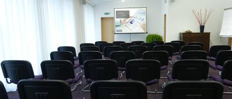 Uma sala de conferências com fileiras de cadeiras, uma tela de projetor e um quadro branco.