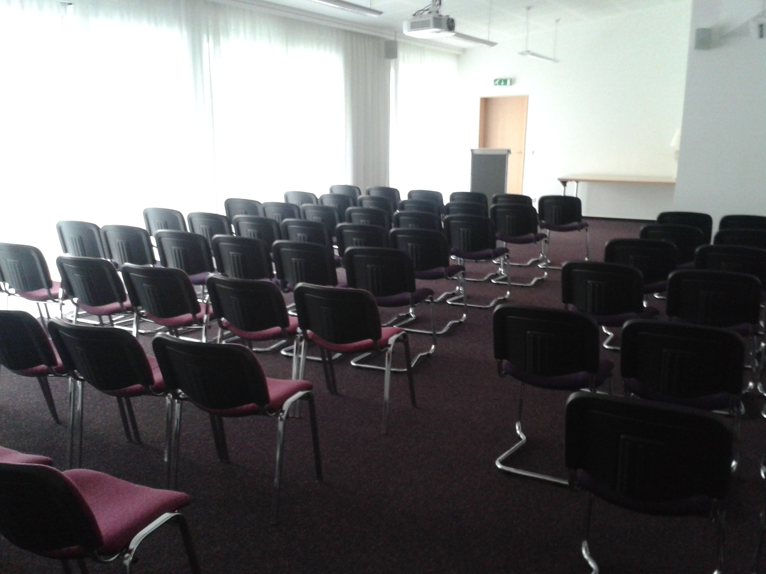 Sala de conferências com fileiras de cadeiras pretas e piso coberto por carpete.