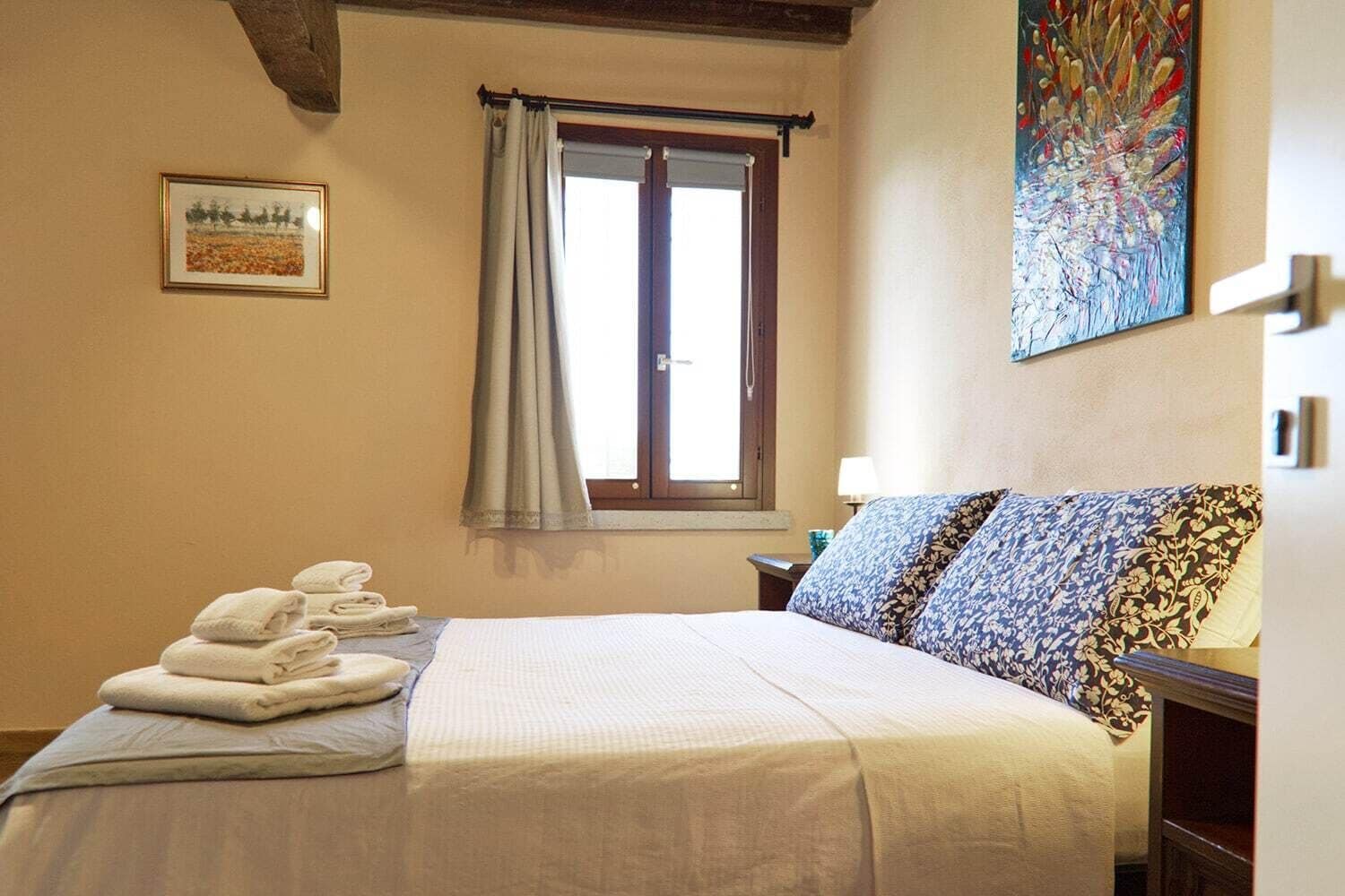 Quarto de hotel com cama, almofadas, janela com cortinas, um quadro na parede e uma fotografia emoldurada.