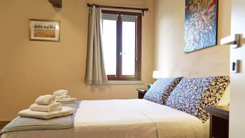 Quarto de hotel com cama, almofadas, janela com cortinas, um quadro na parede e uma fotografia emoldurada.