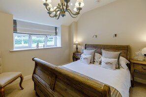 Quarto com cama de madeira, mesa de cabeceira com abajur, janela com cortinas e um lustre.