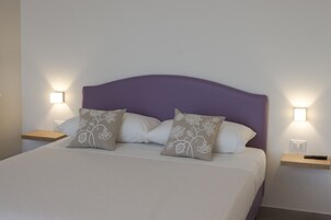 Uma cama bem arrumada com cabeceira roxa, duas mesas de cabeceira com abajures e almofadas estampadas com flores.