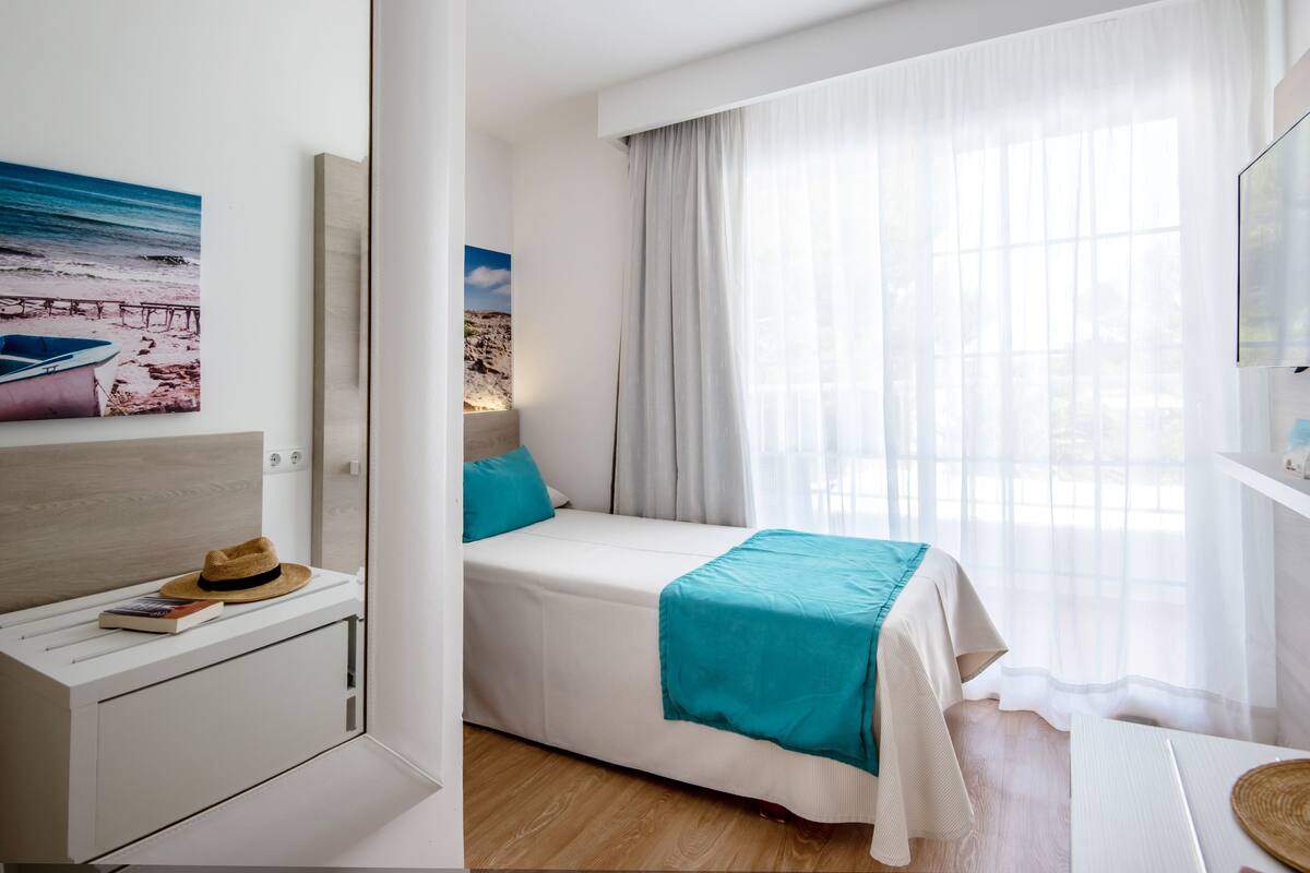 Quarto moderno com cama, mesa de cabeceira com chapéu e livros, janela com cortinas leves e obra de arte na parede.