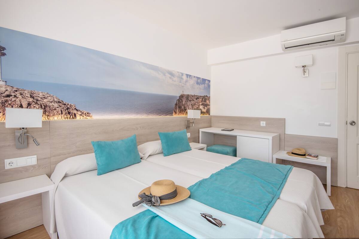 Quarto de hotel com cama, escrivaninha, cadeira e uma grande pintura de uma paisagem costeira.